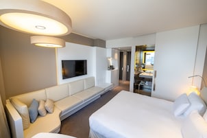 Quarto de hotel moderno com uma cama grande, um sofá, uma TV fixada na parede e um closet espaçoso.