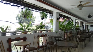Um espaço amplo e interno, com várias mesas e cadeiras, janelas grandes com plantas pendentes e ventiladores de teto.