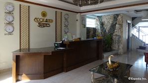 Uma área de recepção de hotel com um balcão de madeira, relógios exibindo horários para Manila, Londres, Sydney, Tóquio, Nova York e Canadá, e uma parede de pedra com uma escada que leva para cima.