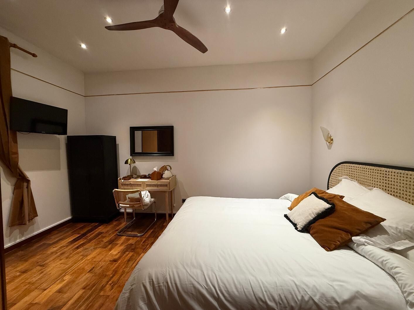 Quarto com cama, escrivaninha com cadeira, televisão e ventoinha no teto.