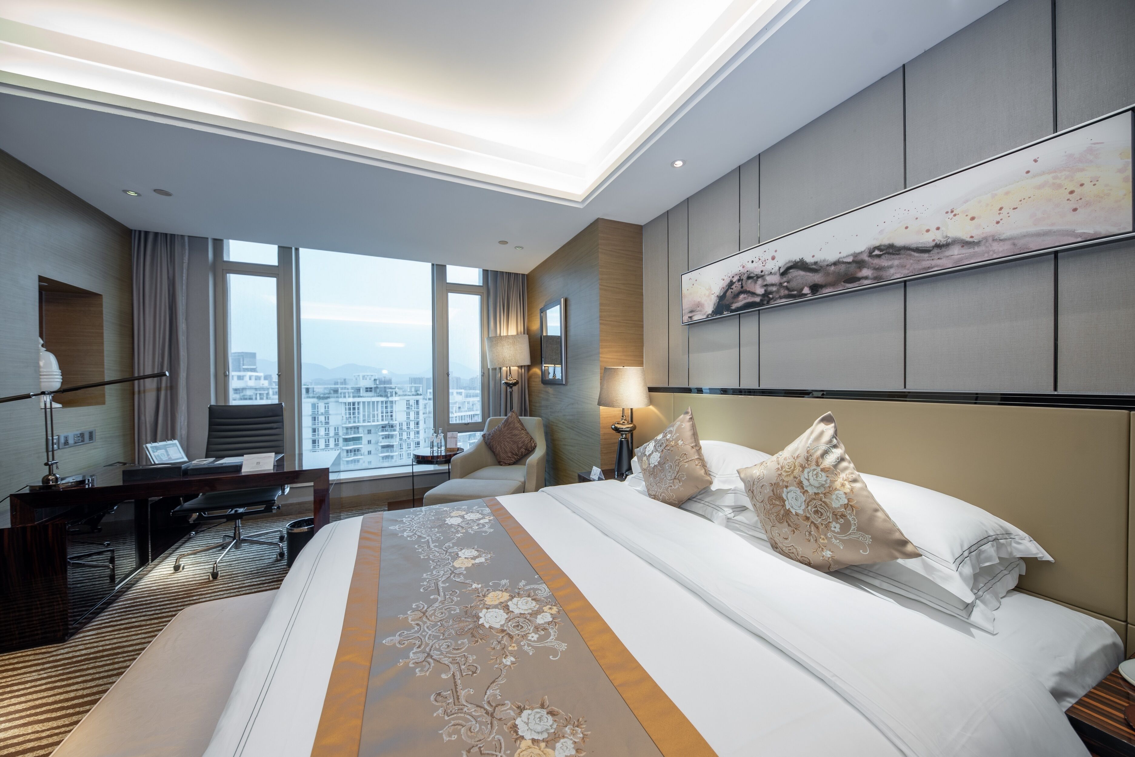Quarto de hotel moderno com uma cama grande, uma escrivaninha e uma cadeira. Há uma janela ampla com vista para a cidade.