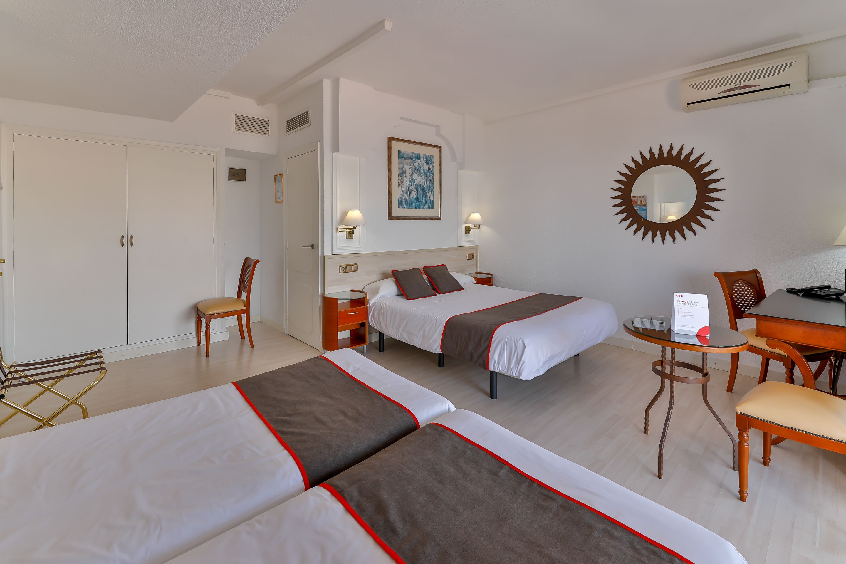 Quarto de hotel com duas camas, uma escrivaninha, uma cadeira, um guarda-roupa e um espelho fixado na parede.