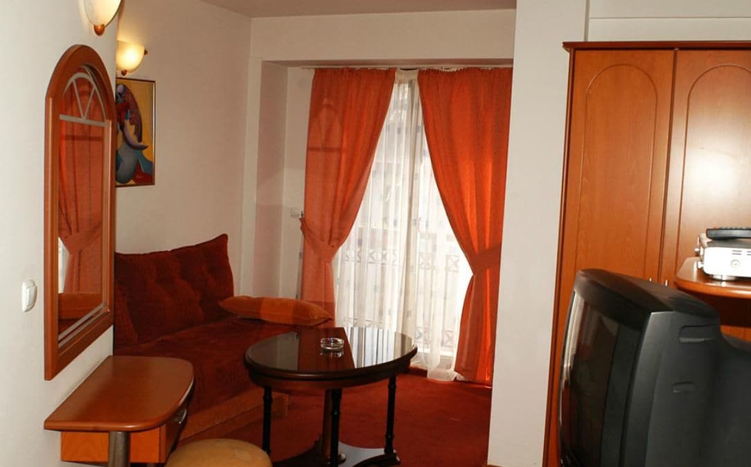 Quarto de hotel compacto com sofá, mesa pequena, televisão e porta que dá para outro espaço.