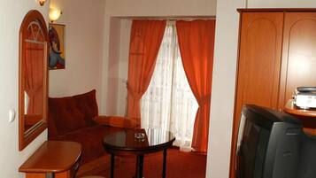 Quarto de hotel compacto com sofá, mesa pequena, televisão e porta que dá para outro espaço.