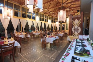 Um restaurante amplo, com várias mesas e cadeiras, grandes janelas e luminárias pendentes.