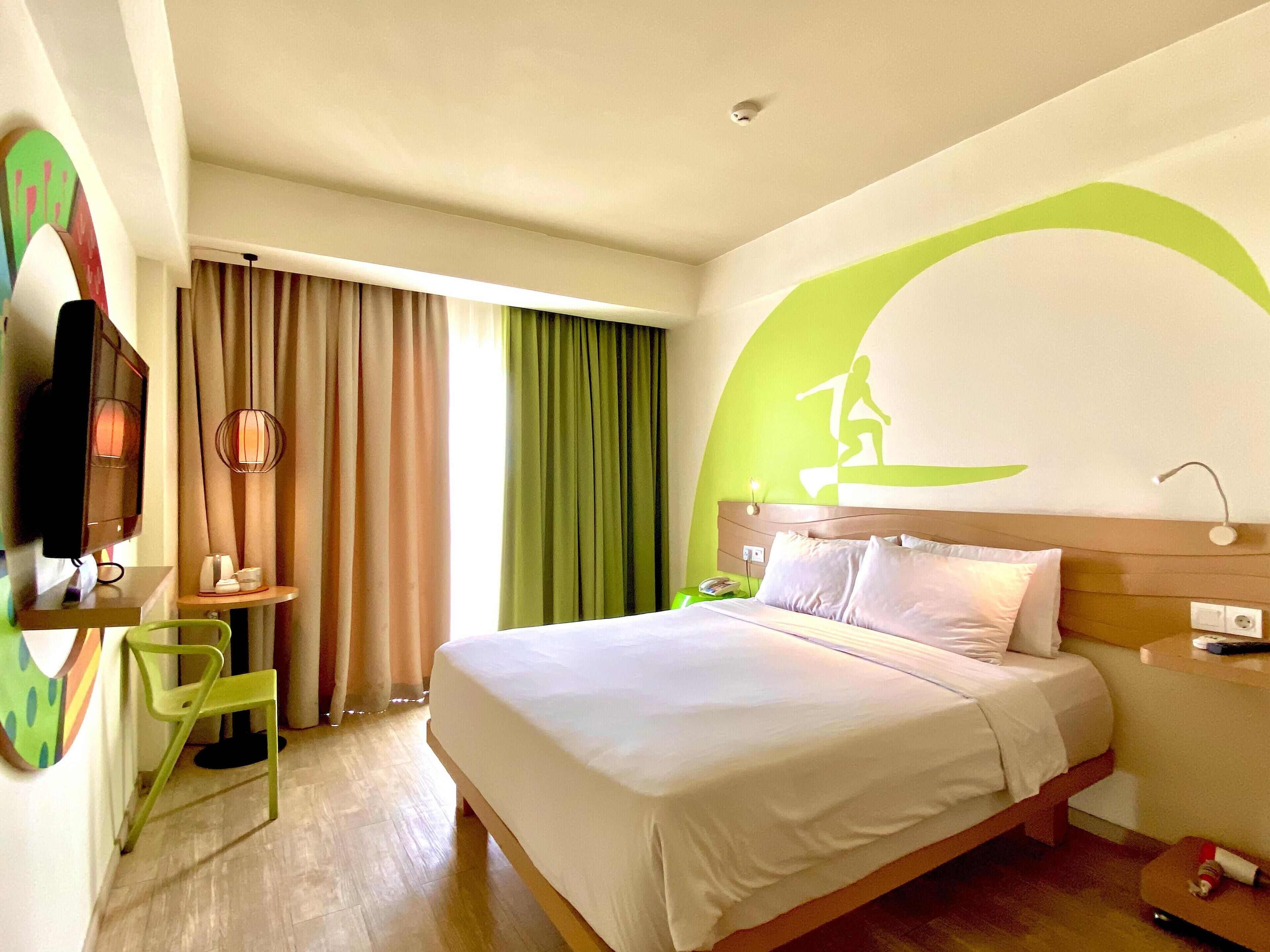 Quarto de hotel com cama, escrivaninha, cadeira, televisão e uma janela grande com cortinas.