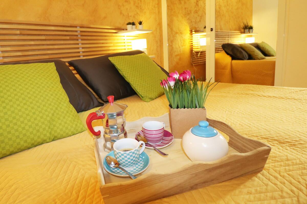 Cama bem arrumada com uma bandeja de itens de café da manhã, incluindo chaleira, xícaras e um pequeno vaso com flores.
