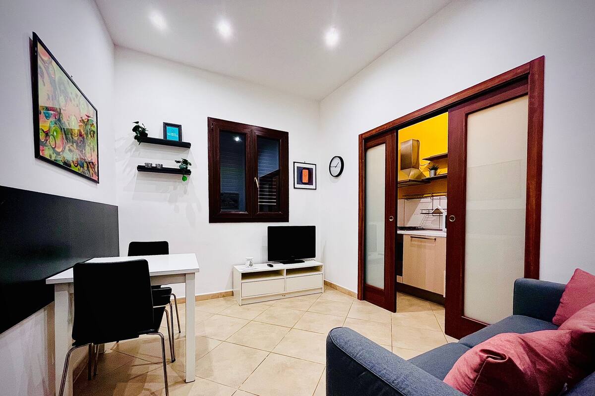 Uma sala moderna com área de jantar, televisão e cozinha visível através de uma porta aberta.