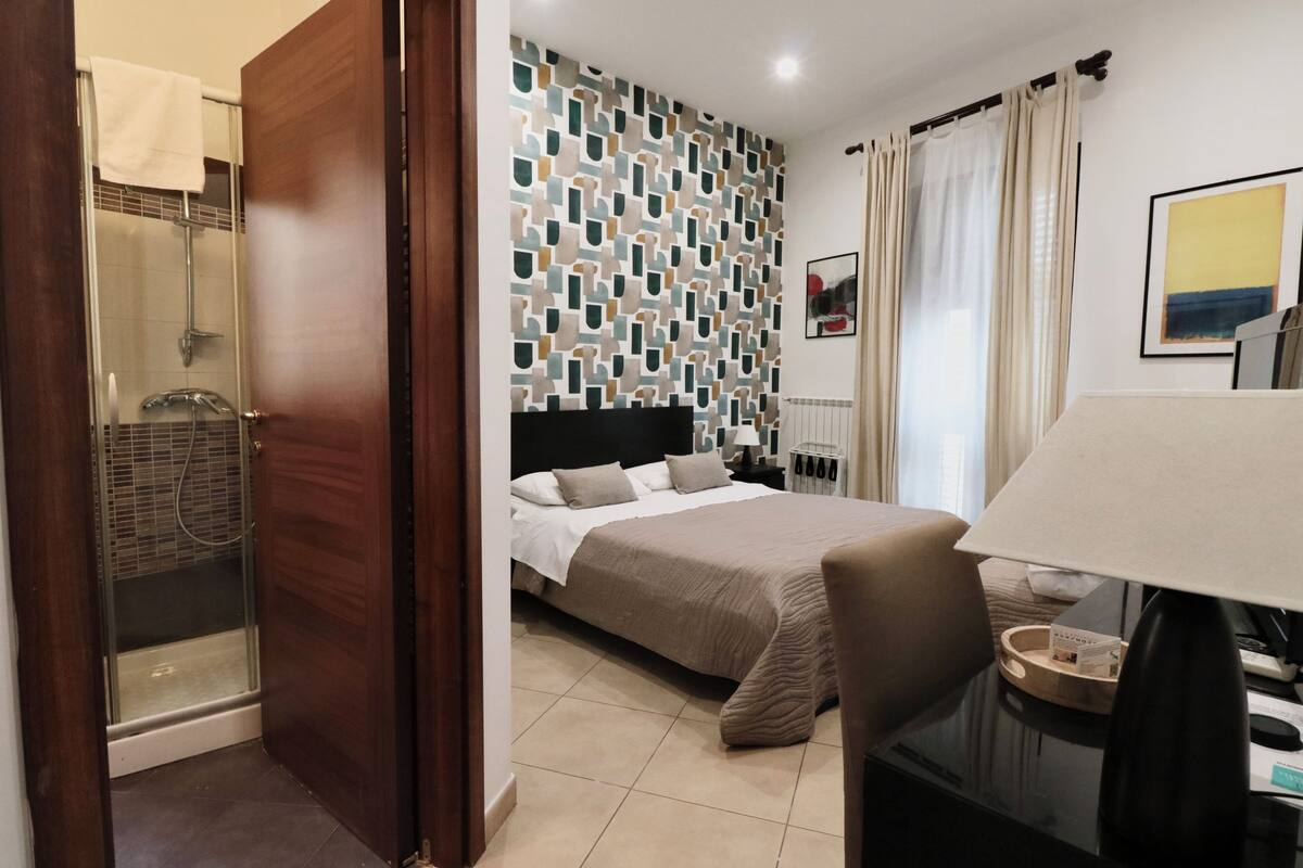 Quarto de hotel com chuveiro, cama com cabeceira estampada, escrivaninha com abajur e janela com cortinas.
