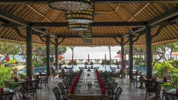 Uma área de refeições ao ar livre com uma mesa comprida e cadeiras, com vista para uma piscina e para o mar.