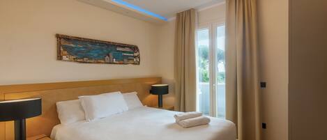 Quarto de hotel com uma cama, duas mesas de cabeceira com luminárias, um quadro na parede e uma janela com cortinas.
