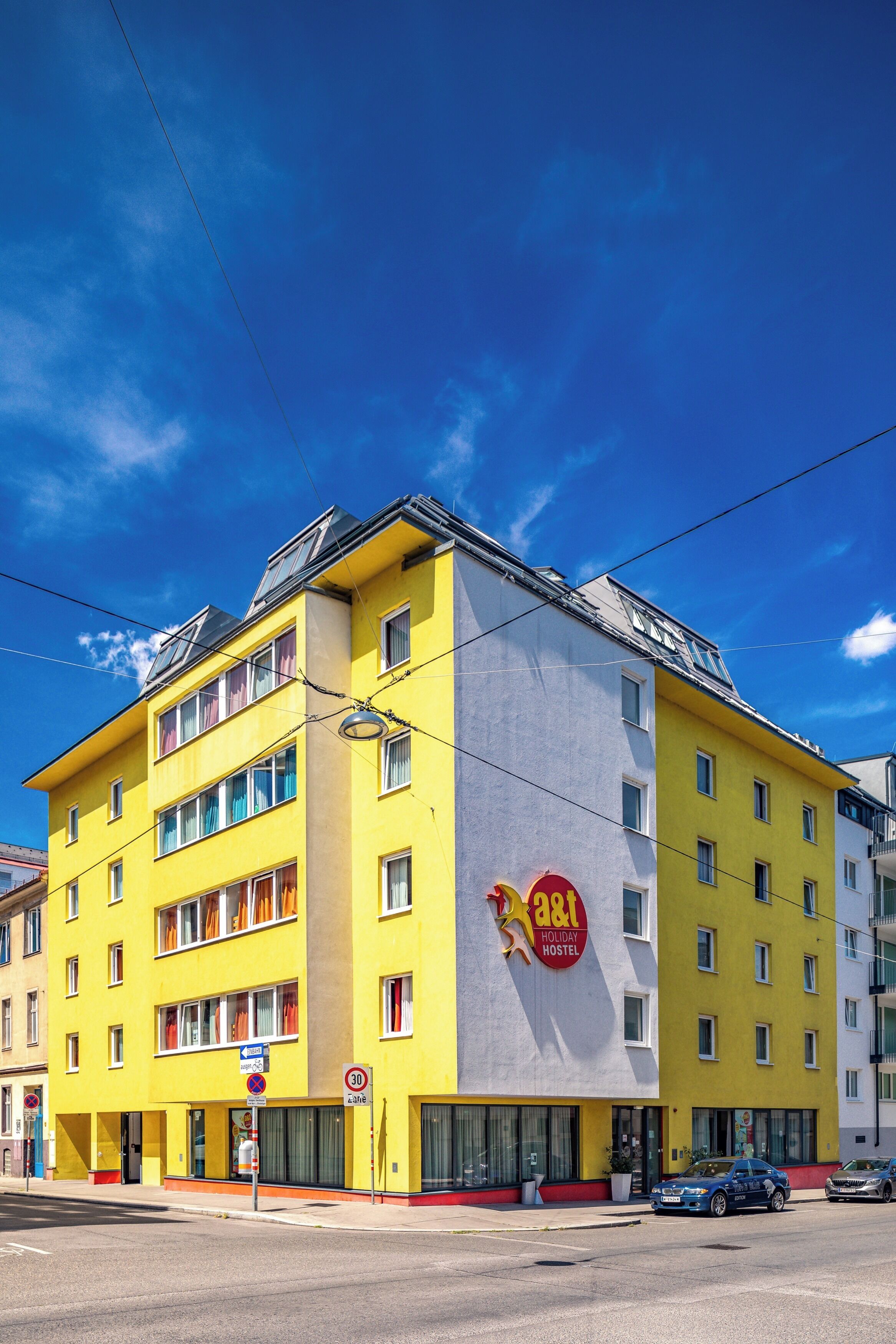 Um edifício de vários andares com fachada amarela, logotipo visível do 'A&J Holiday Hostel' e placa de limite de velocidade indicando '30'.