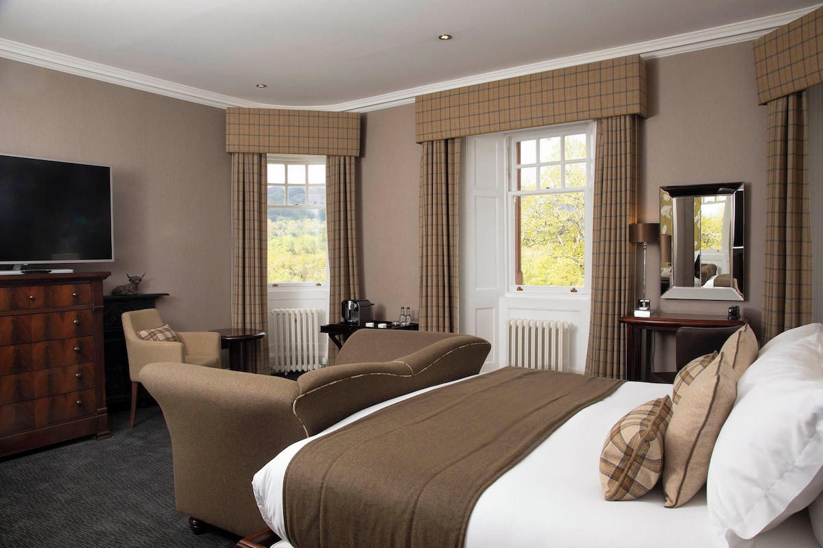 Quarto de hotel com cama, televisão, cômoda, cadeira e escrivaninha com abajur.