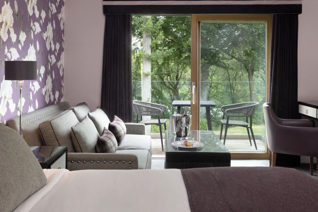 Quarto de hotel com uma cama, um sofá, uma mesa de jantar com cadeiras e vista para o exterior através de uma porta de vidro.