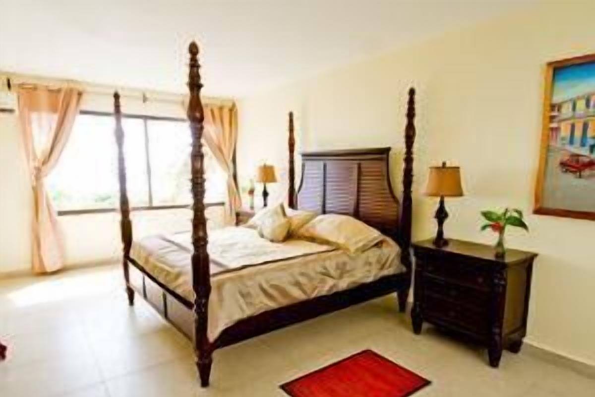 Uma cama com dossel, cabeceira de madeira, mesa de cabeceira com abajur e tapete vermelho sobre piso de azulejo.