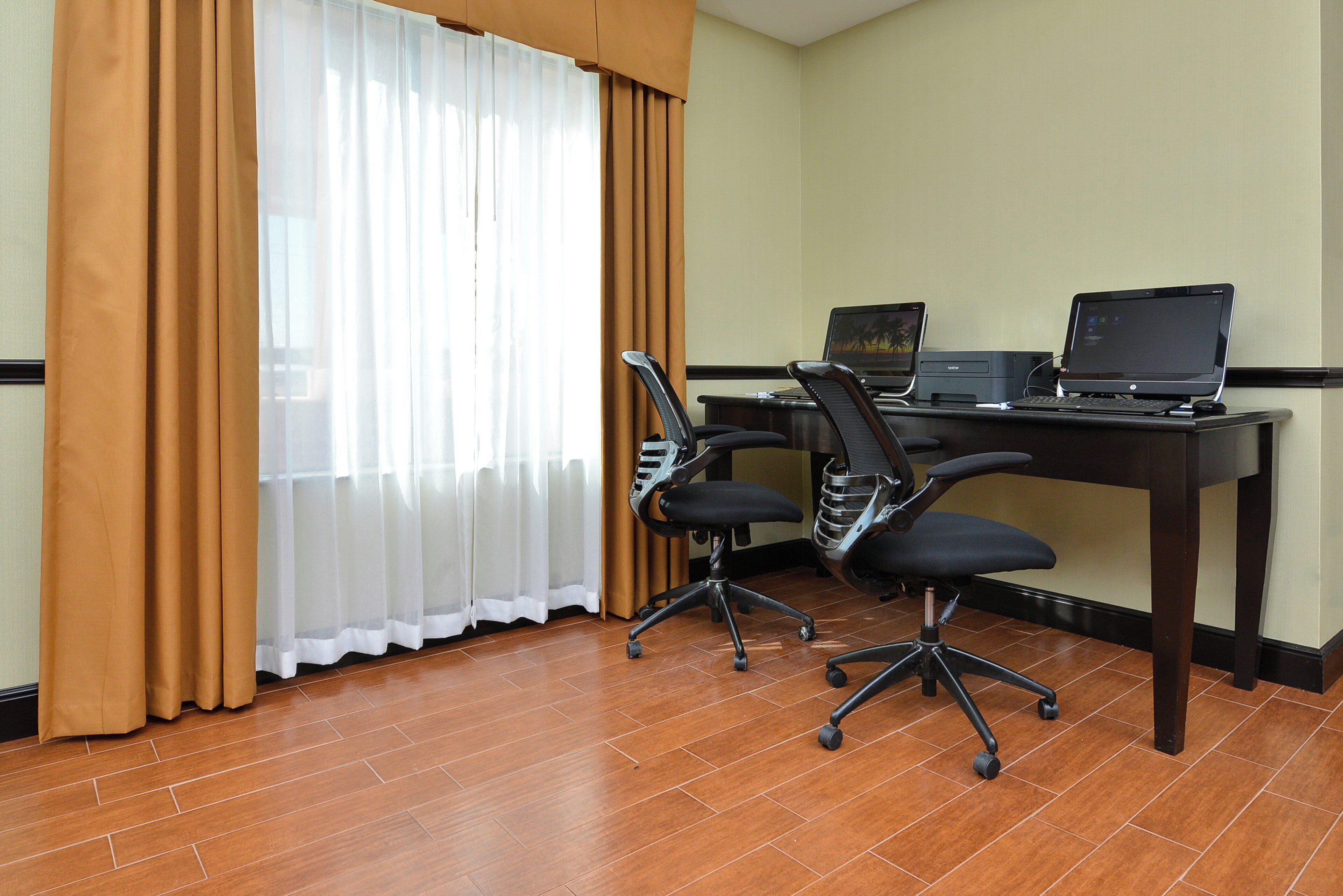 Quarto de hotel com piso de madeira, uma escrivaninha com dois laptops, duas cadeiras de escritório e uma janela com cortinas leves.