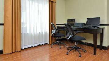 Quarto de hotel com piso de madeira, uma escrivaninha com dois laptops, duas cadeiras de escritório e uma janela com cortinas leves.