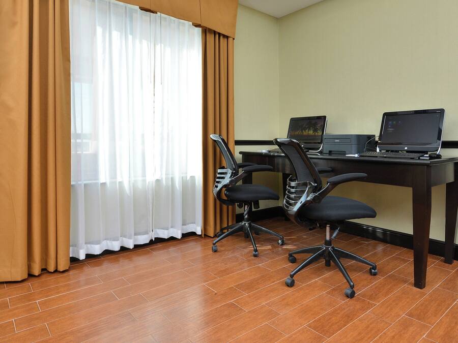 Quarto de hotel com piso de madeira, uma escrivaninha com dois laptops, duas cadeiras de escritório e uma janela com cortinas leves.