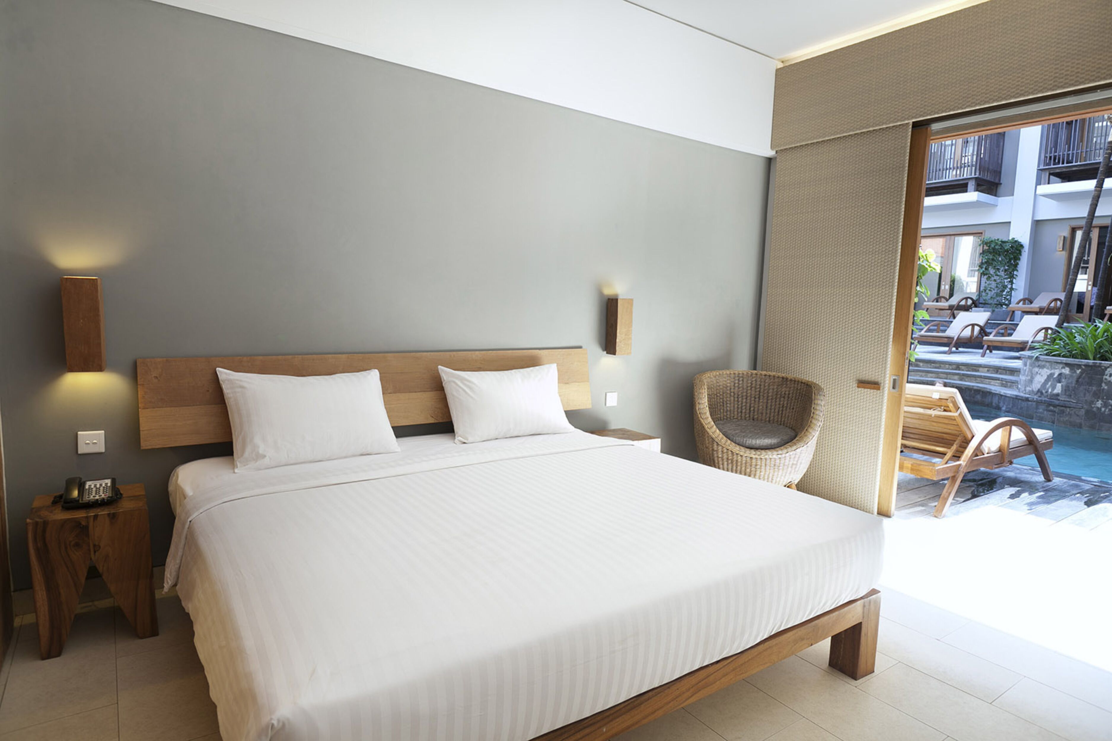 Quarto de hotel moderno com uma cama grande, uma mesa de cabeceira com telefone, uma poltrona de vime e vista para uma área de piscina.