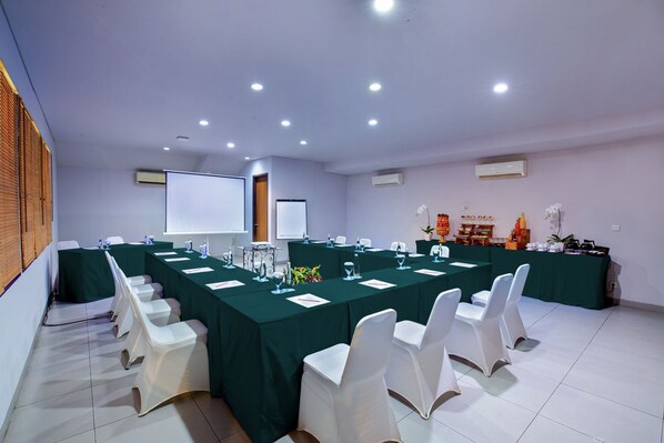 Uma sala de conferências com uma mesa comprida, cadeiras brancas e uma tela de projetor.