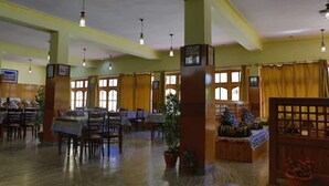 Um hall amplo com piso de madeira, várias mesas e cadeiras, e diversos vasos de plantas.