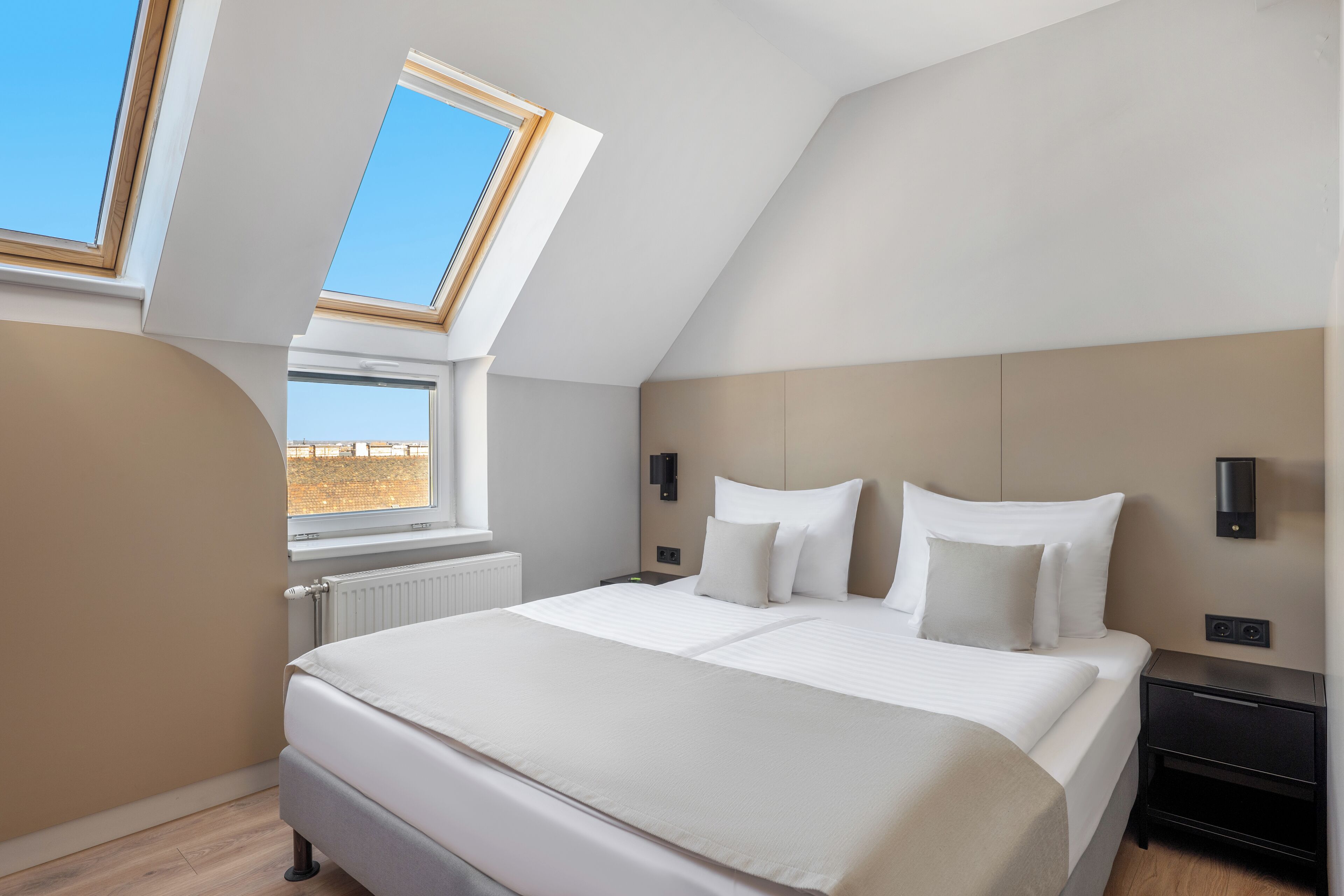 Quarto com claraboia, cama com roupa branca, mesa de cabeceira e luminária na parede.