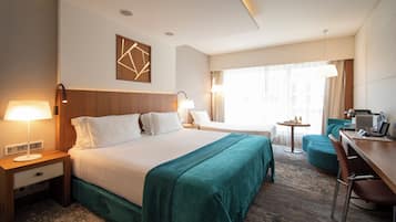 Quarto de hotel com uma cama grande, duas mesas de cabeceira, uma escrivaninha com cadeira, uma janela com cortinas e uma obra de arte pendurada na parede.