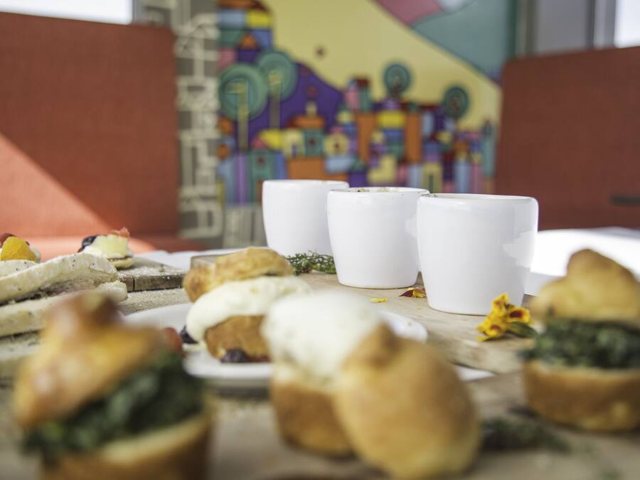 Uma mesa com diversos alimentos e copos brancos, provavelmente para bebidas, em um ambiente com obras de arte coloridas ao fundo.