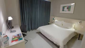 Quarto de hotel com cama, escrivaninha, cadeira, abajur e um quadro na parede.