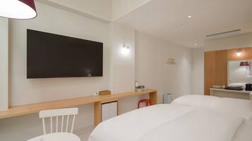 Quarto de hotel com uma televisão de tela plana grande presa na parede, uma escrivaninha de madeira com cadeira, uma pequena geladeira e duas camas.