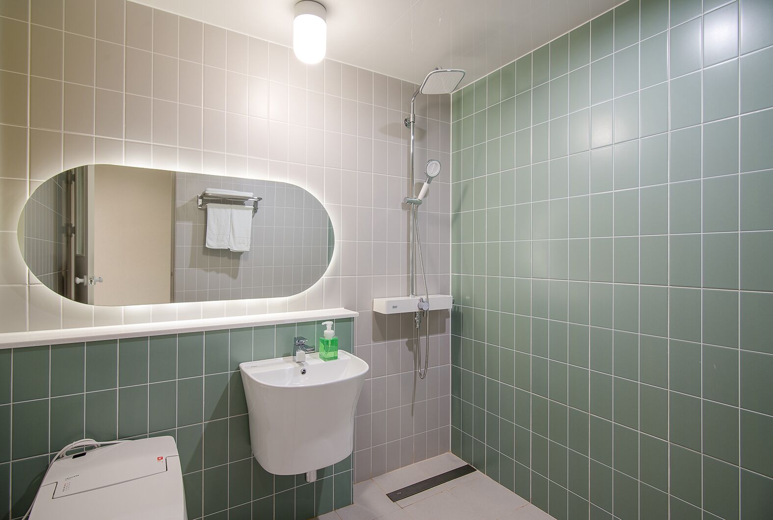Casa de banho com uma pia branca, azulejos verdes, chuveiro e um espelho com uma toalha pendurada.