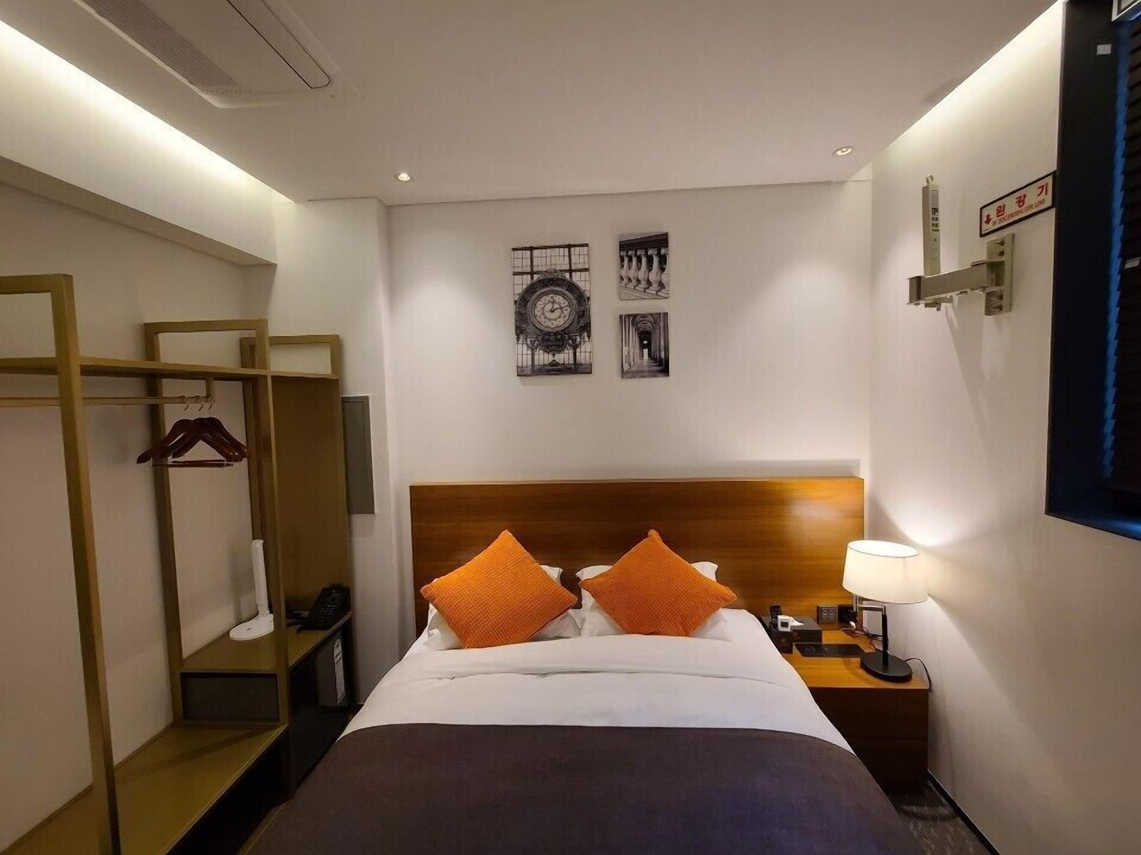 Quarto de hotel com cabeceira de madeira, dois travesseiros laranja, mesa de cabeceira com abajur e guarda-roupa com espelho.
