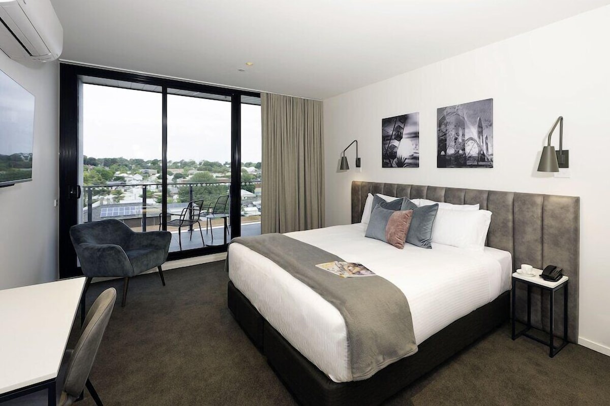 Quarto de hotel com uma cama grande, uma escrivaninha, uma cadeira, varanda com vista e obras de arte nas paredes.