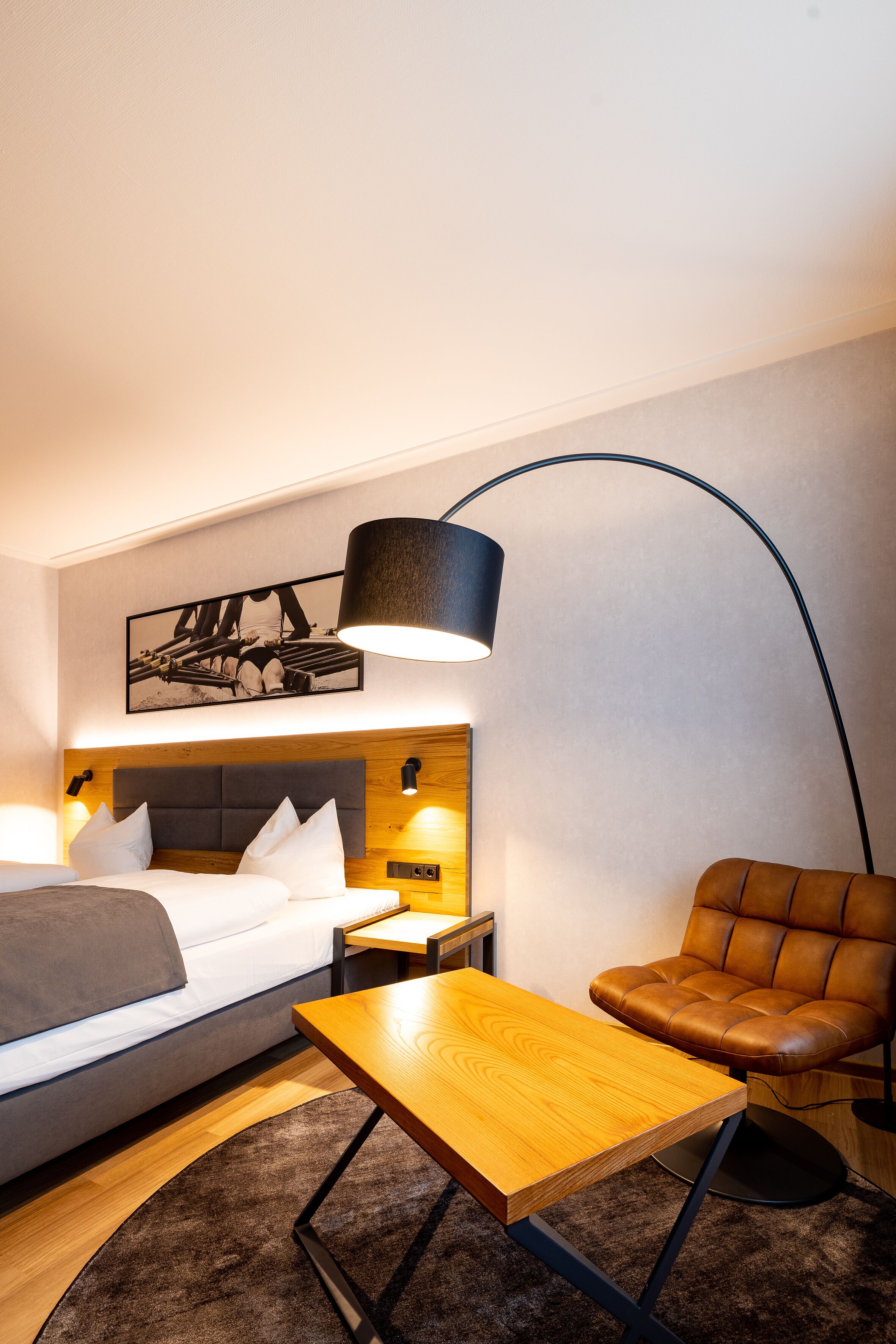 Quarto de hotel moderno com uma cama, uma mesa de madeira, uma poltrona de couro, um abajur e uma obra de arte na parede.