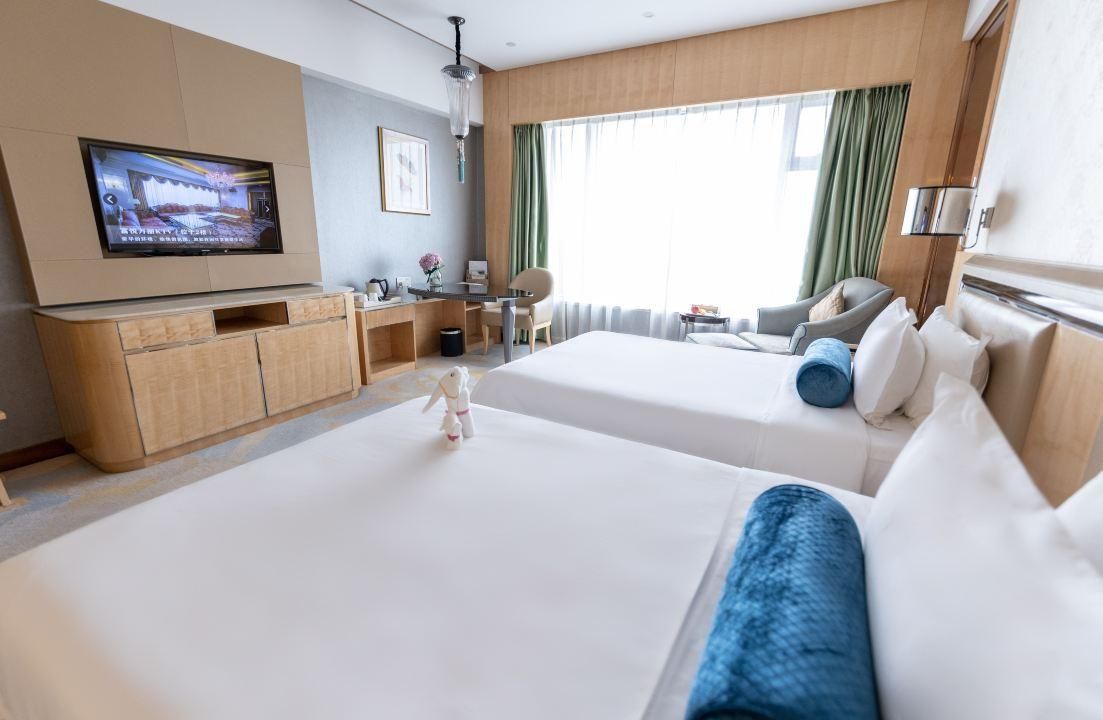 Quarto de hotel com duas camas, uma escrivaninha, televisão e uma janela grande com cortinas.