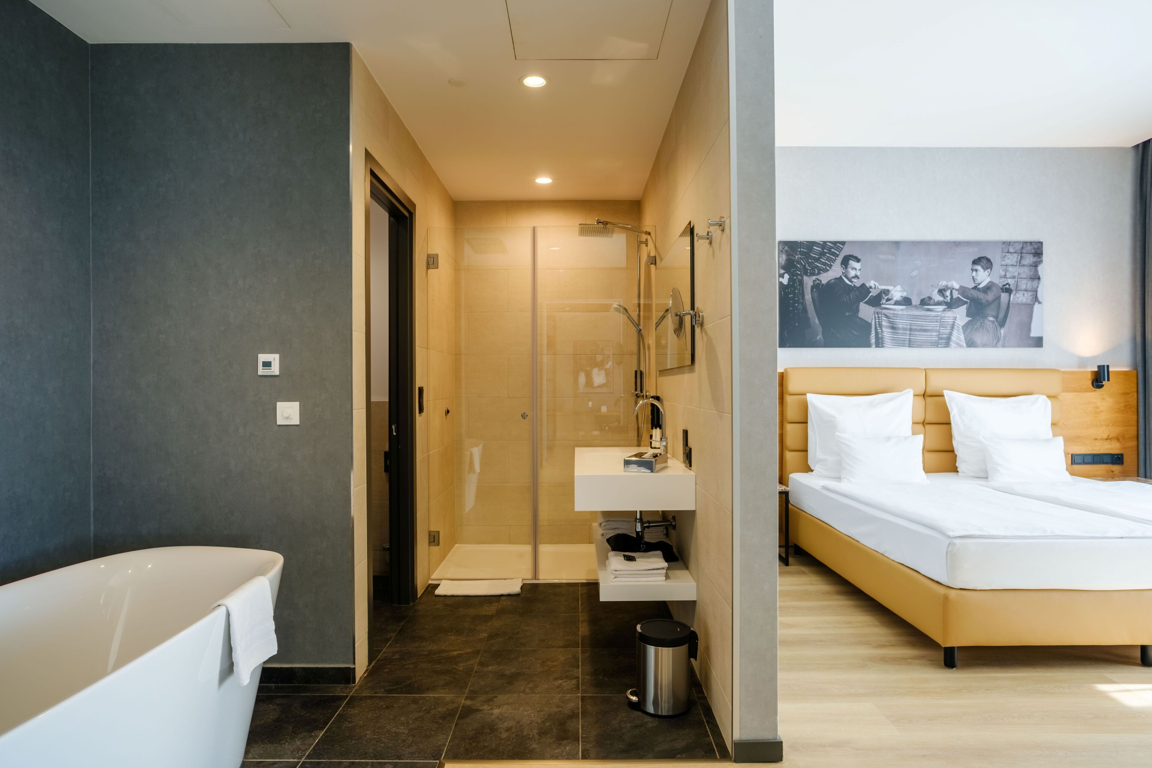 Quarto de hotel moderno com banheira, chuveiro, cama, mesa de cabeceira e espelho fixado na parede.
