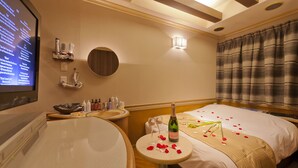 Quarto de hotel com uma banheira grande, um espelho redondo, mesa de cabeceira com champanhe e taças, e uma cama com pétalas de rosa.