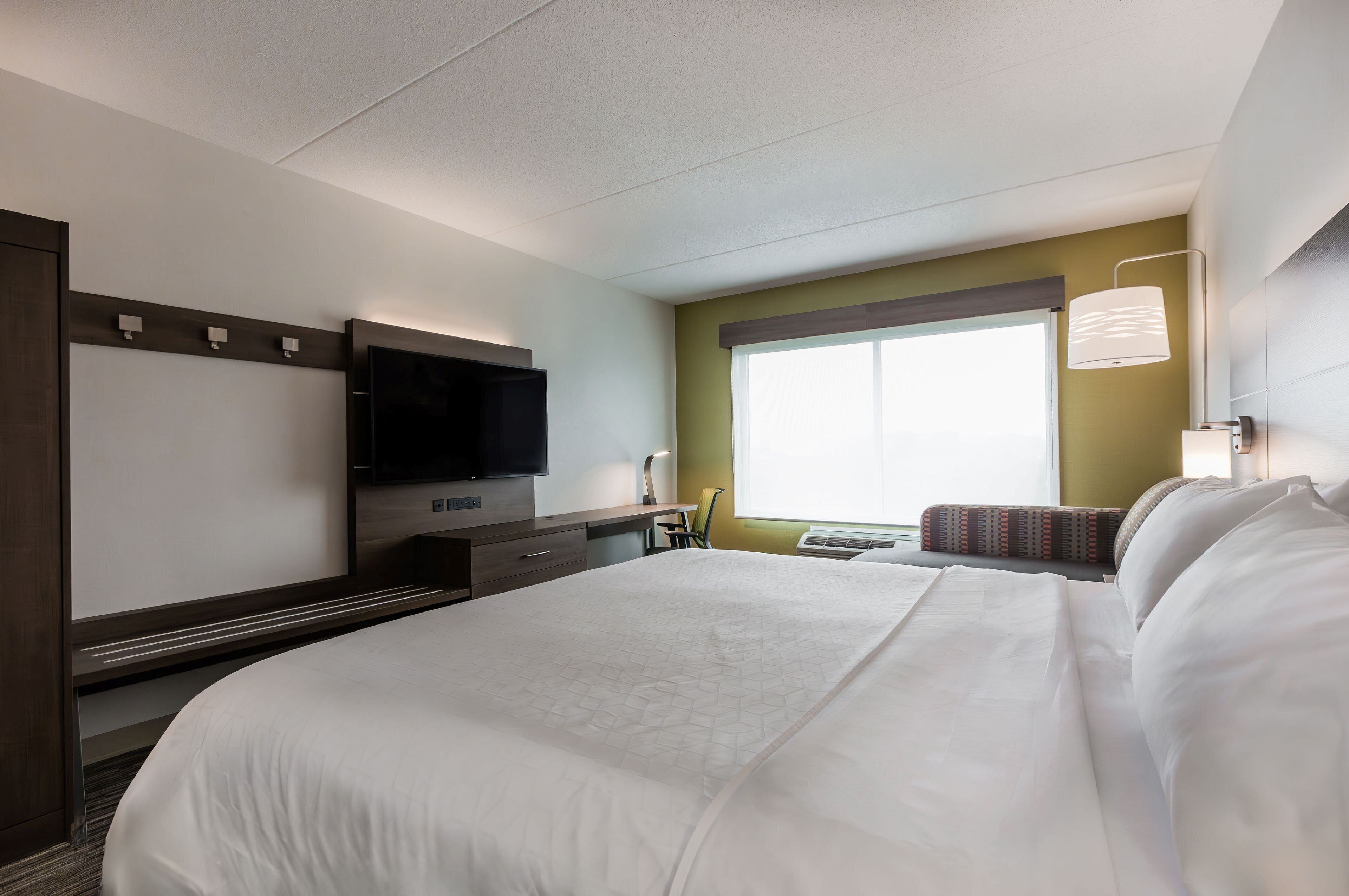 Quarto de hotel com uma cama grande, uma mesa com televisão de tela plana, uma cadeira e uma janela com persianas.