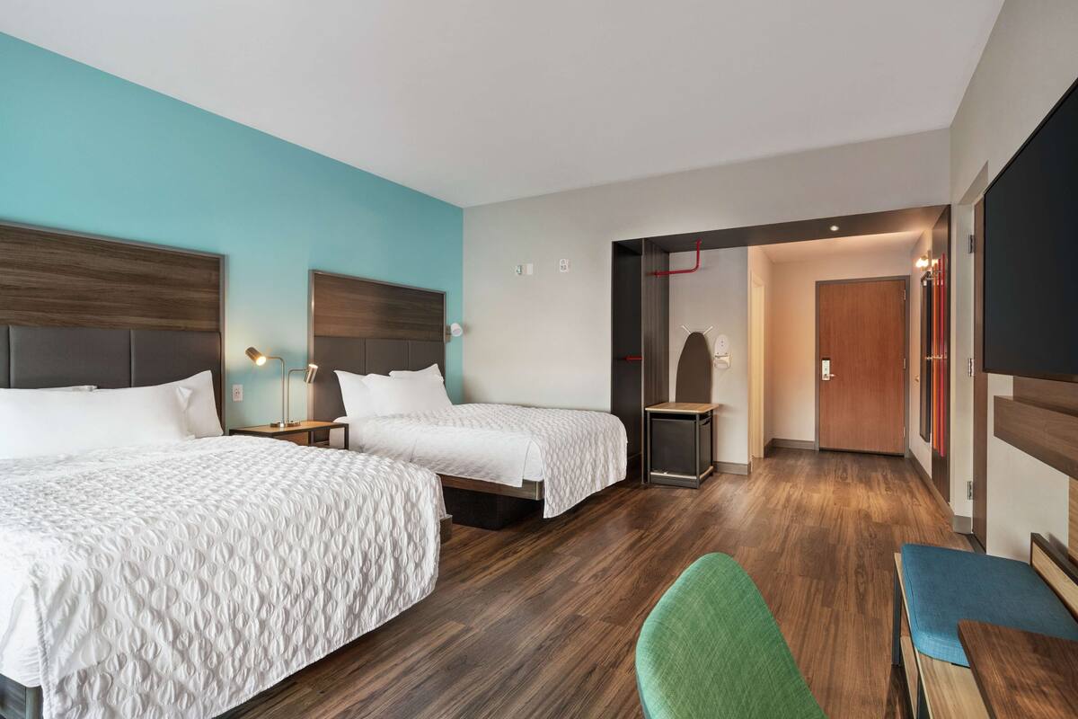 Quarto de hotel com duas camas, uma pequena mesa, uma cadeira e uma porta que dá para um corredor.