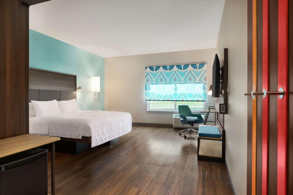 Quarto de hotel com cama, escrivaninha, cadeira, televisão e janela com persianas.