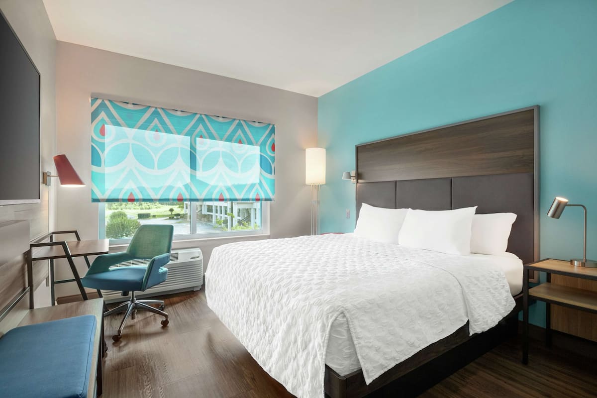 Quarto de hotel com uma cama grande, uma escrivaninha, uma cadeira, uma janela com cortinas estampadas e vista para o exterior.