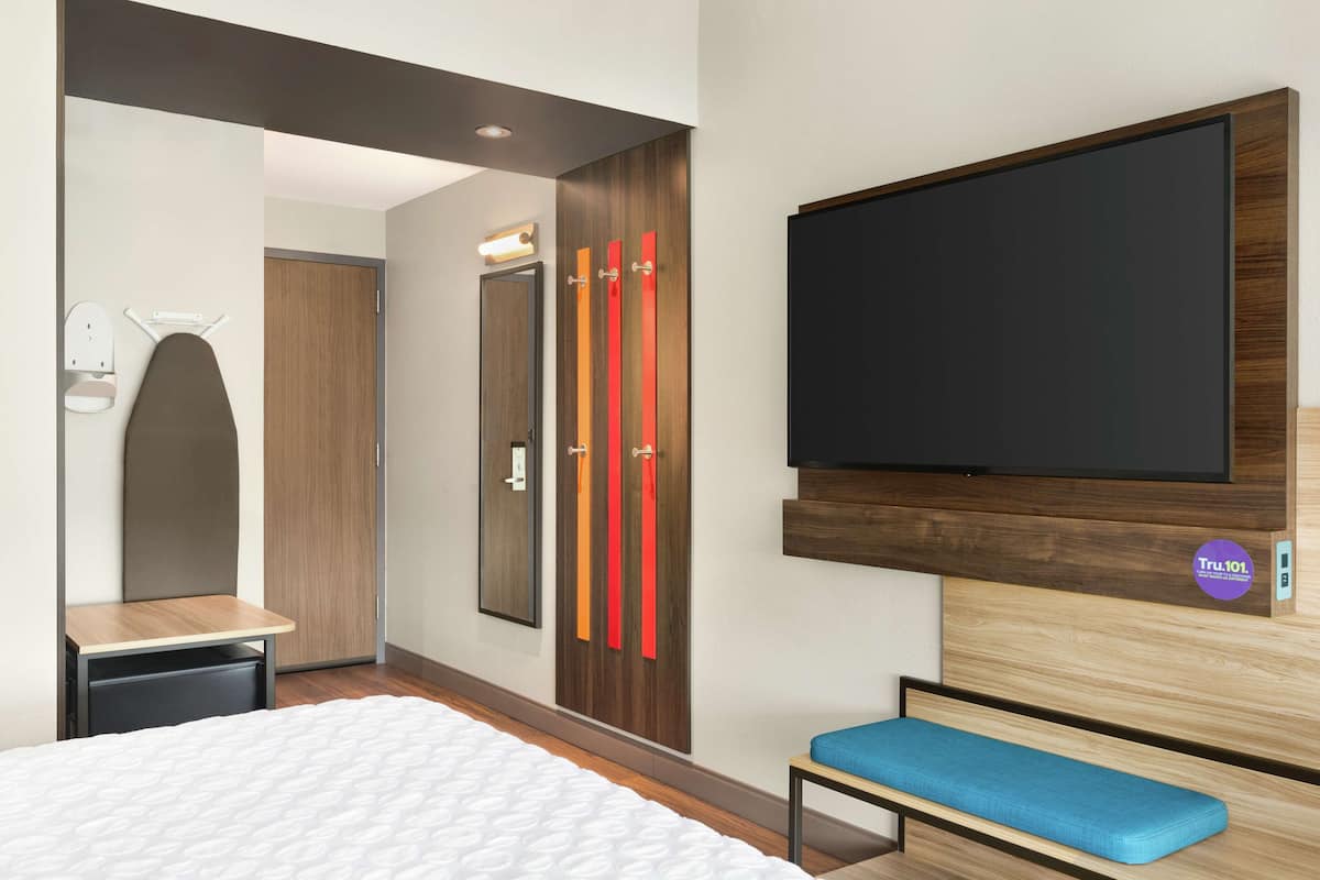 Quarto de hotel com cama, um banquinho, televisão, espelho e guarda-roupa.