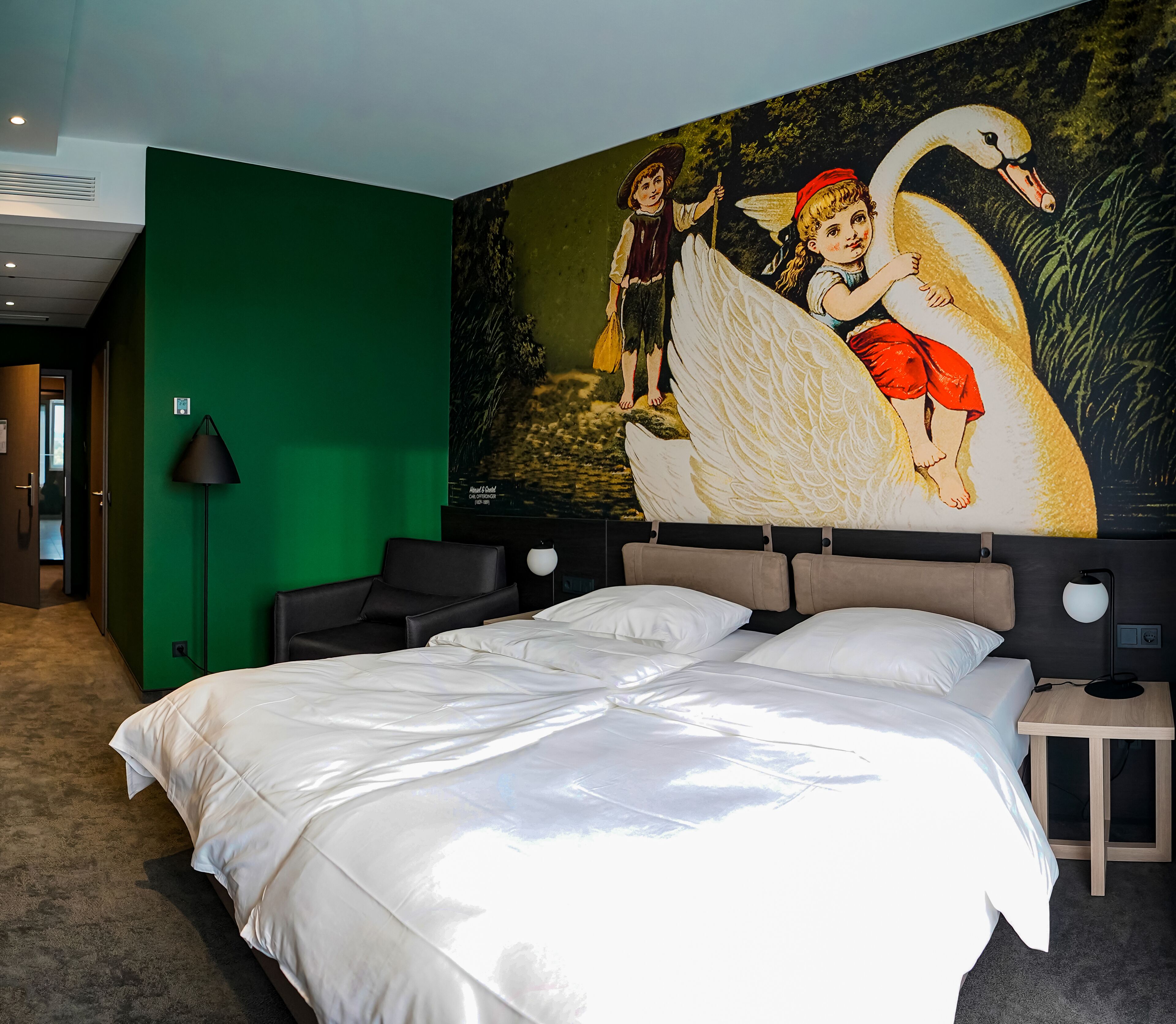 Quarto de hotel com uma cama grande, uma parede de destaque verde e um mural com cisne e crianças.