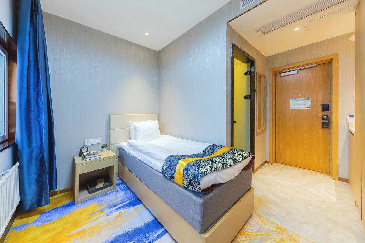 Quarto de hotel com cama, mesa de cabeceira, pequena mesa, cortina azul e porta de madeira.