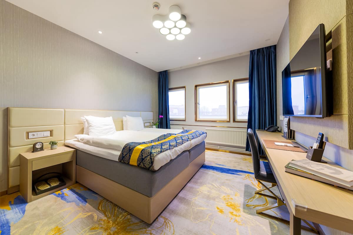 Quarto de hotel com cama, escrivaninha, cadeira, televisão e janela com cortinas.