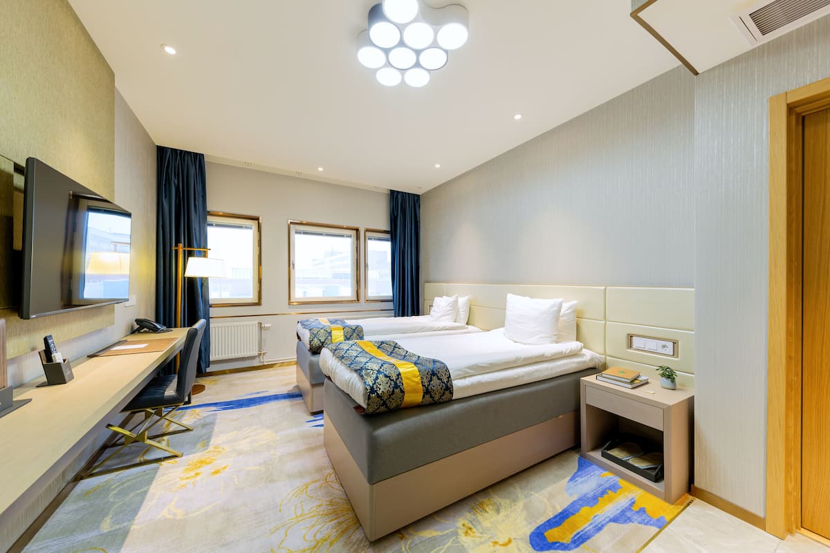 Quarto de hotel com uma cama grande, uma escrivaninha com cadeira, televisão e mesa de cabeceira com abajur.