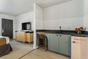 Cozinha compacta com geladeira embutida, área de jantar com uma cadeira e uma cama no quarto adjacente.