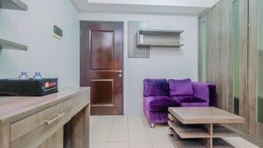 Quarto compacto com uma escrivaninha de madeira, um sofá roxo, uma mesa pequena, uma porta e uma prateleira fixada na parede.