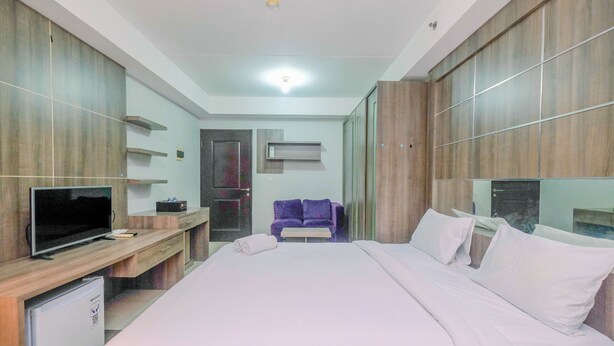 Quarto de hotel com uma cama, televisão, escrivaninha, mini-geladeira e sofá roxo.
