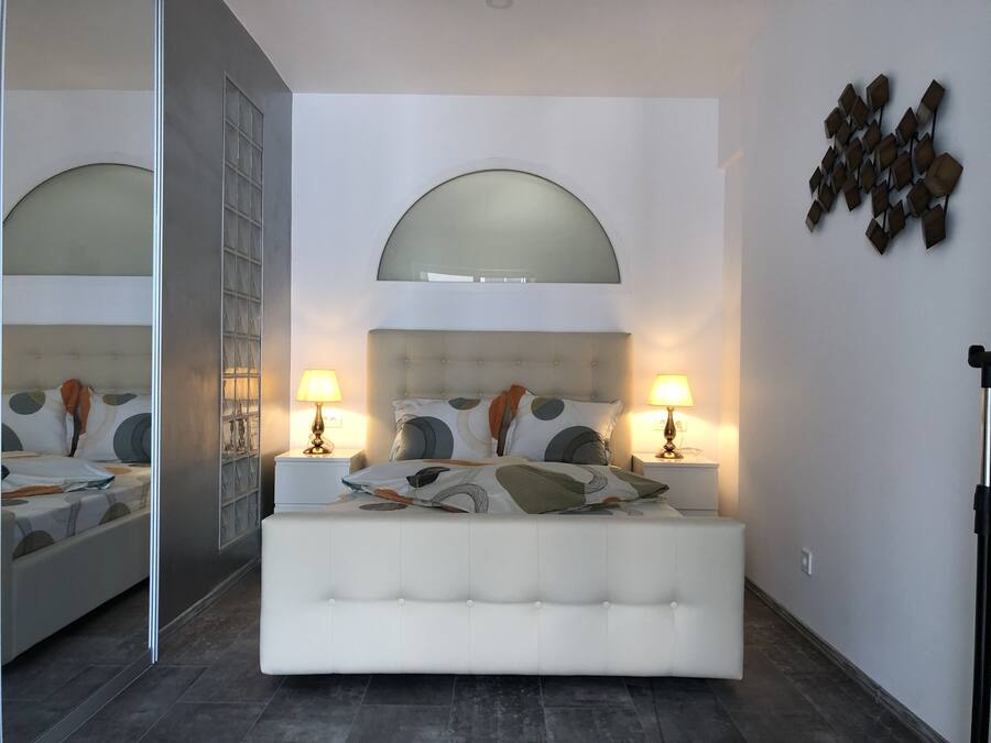 Um quarto moderno com uma cama grande, duas mesas de cabeceira com abajures, um espelho grande e um detalhe decorativo na parede.